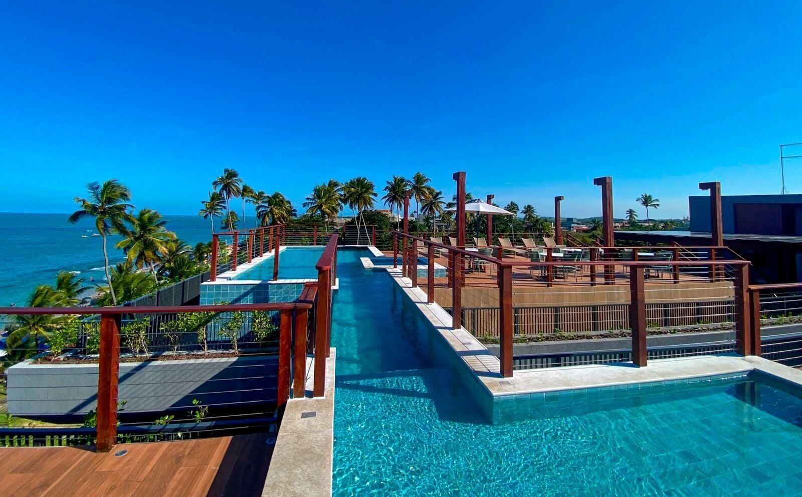 Área de piscina com água azul cristalina, deck de madeira e palmeiras, com vista para o oceano.