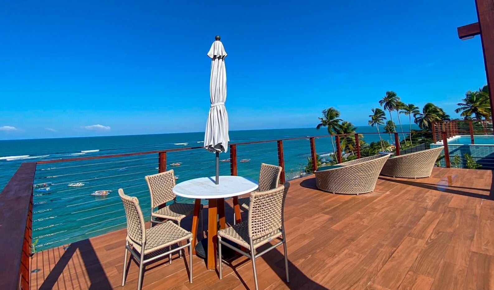 Um deck de madeira com móveis de vime e uma mesa, com vista para um mar azul e límpido, palmeiras e a vastidão do oceano.