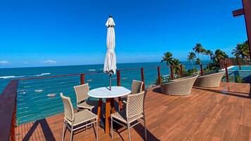 Um deck de madeira com móveis de vime e uma mesa, com vista para um mar azul e límpido, palmeiras e a vastidão do oceano.