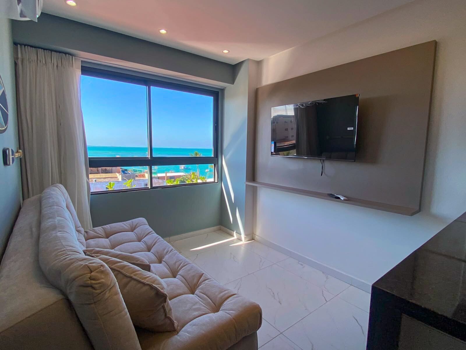 Uma sala de estar moderna com uma janela grande, uma TV de tela plana na parede e um sofá bege.