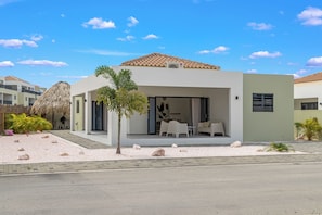 Uma casa moderna de dois andares com telhado plano, um pátio com móveis de exterior e um pequeno jardim com pedras e plantas.