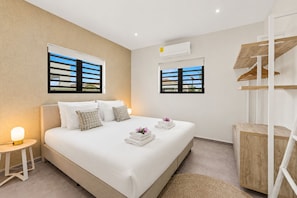 Quarto de hotel moderno com uma cama grande, duas janelas com cortinas e um ar condicionado, além de uma prateleira de madeira.