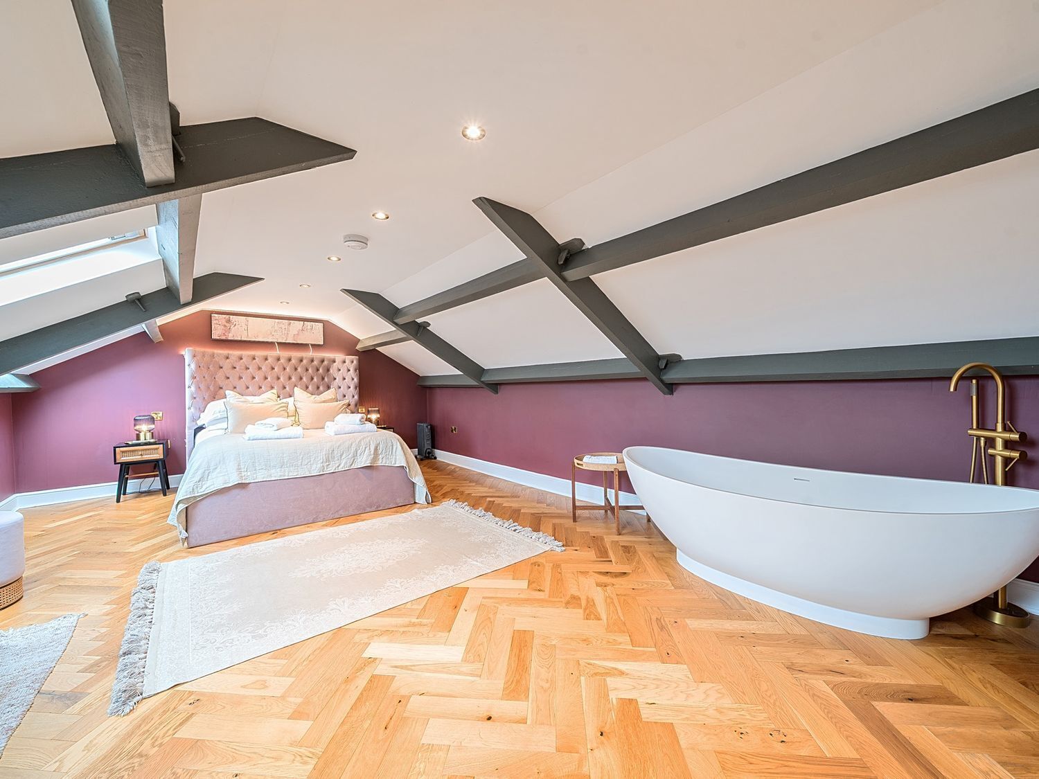 Um quarto com uma cama grande, uma banheira branca e um chão de madeira com padrão herringbone.