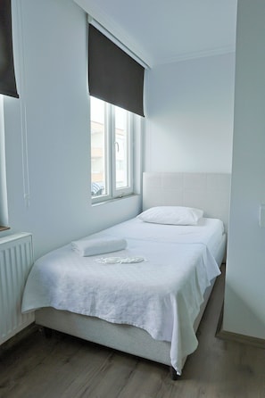 Quarto individual com uma cama, janela, radiador e cabeceira branca.