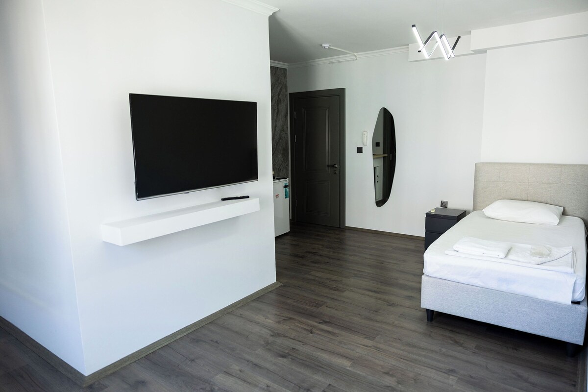 Quarto de hotel moderno com televisão de tela plana fixada em uma prateleira branca, cama com cabeceira cinza, espelho redondo na parede e piso de madeira.