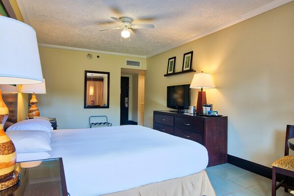 Quarto de hotel com uma cama, televisão, escrivaninha com abajur, poltrona e ventoinha no teto.