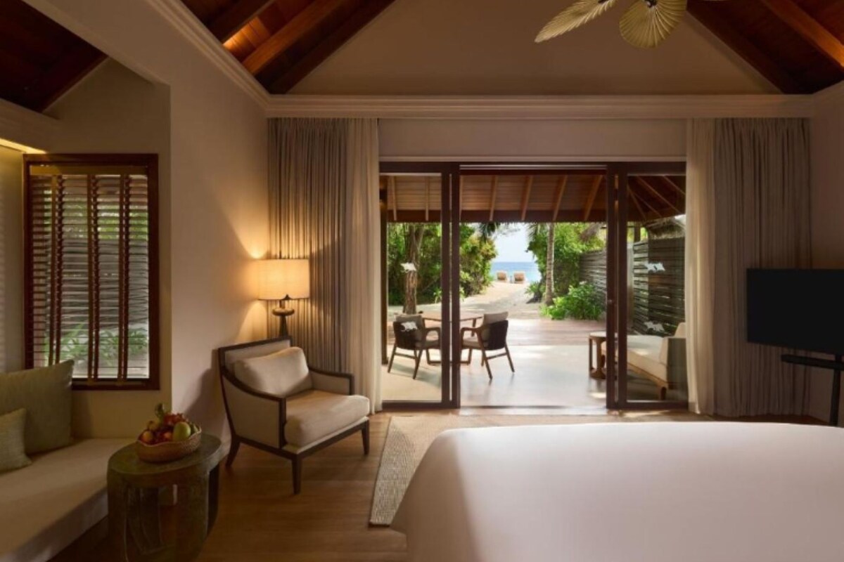 Quarto de hotel com cama, uma cadeira, uma mesinha com frutas, televisão e vista para área externa com mesa e cadeiras.