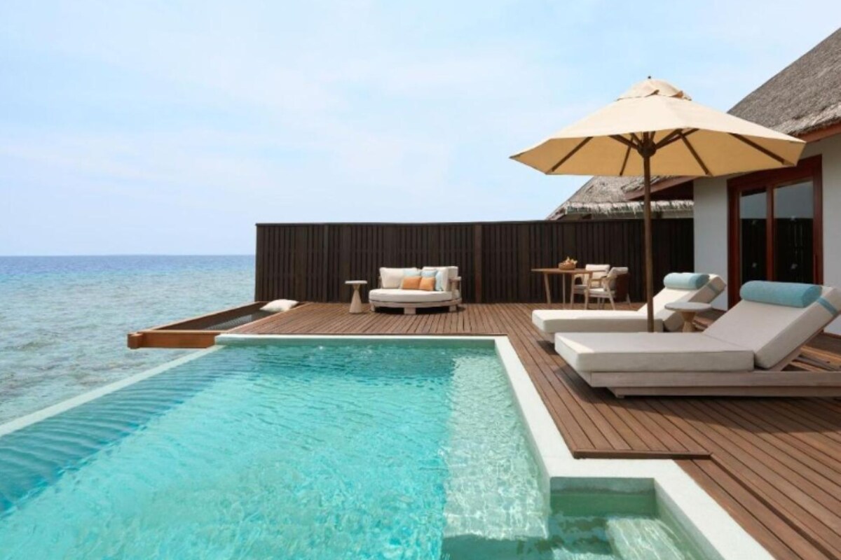 Uma piscina infinita com deck de madeira, espreguiçadeiras e uma mesa com toldo.