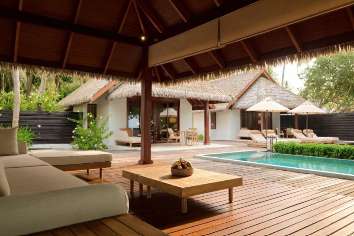 Uma área de piscina ao ar livre com deck de madeira, espreguiçadeiras e uma estrutura com telhado de palha.