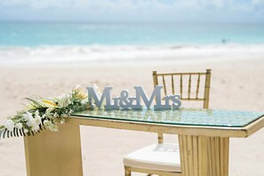 Uma mesa com placa 'Mr & Mrs', uma cadeira e um arranjo floral em um ambiente de praia.