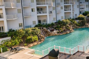 Piscina rodeada por área ajardinada com borda rochosa, deck de madeira e espreguiçadeiras.