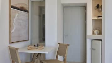 Um quarto pequeno com uma mesa branca, duas cadeiras de vime, uma porta e um frigorífico branco.