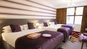 Quarto de hotel com duas camas, uma janela com cortinas e uma pequena mesa com brinquedos e livros.