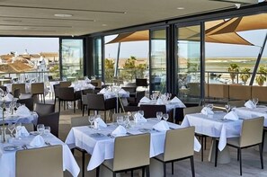 Um restaurante com mesas postas para refeições, grandes janelas e vista para a paisagem.