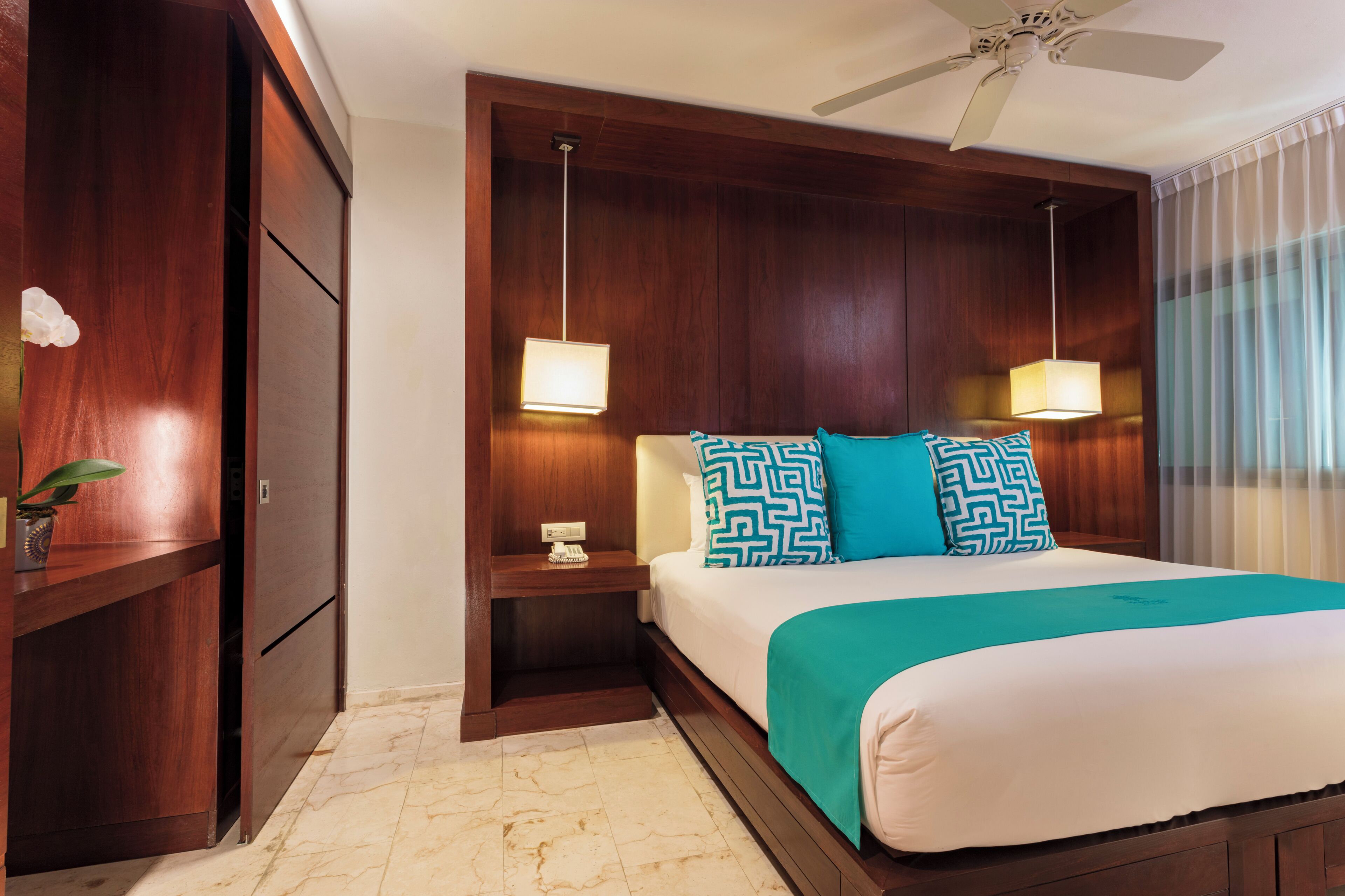 Quarto de hotel com cama, mesas de cabeceira, ventoinha no teto e janela com cortinas.