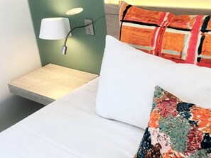 Uma cama com roupa de cama branca, um travesseiro colorido, uma luminária de cabeceira e uma pequena mesa de cabeceira de madeira.