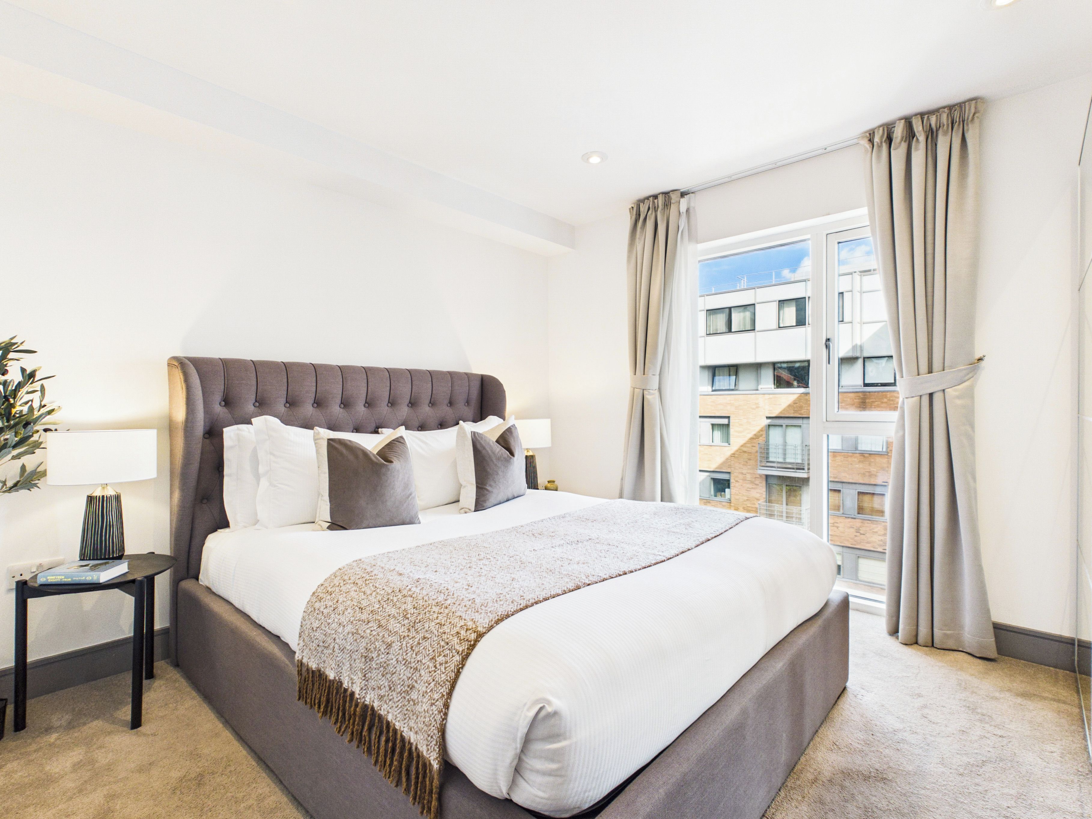 Quarto de hotel com cama grande, mesa de cabeceira, abajur e vista para prédios pela janela.