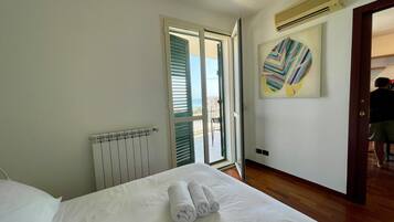 Quarto de hotel com cama, radiador, janela com persianas, porta e parede com uma pintura abstrata colorida.