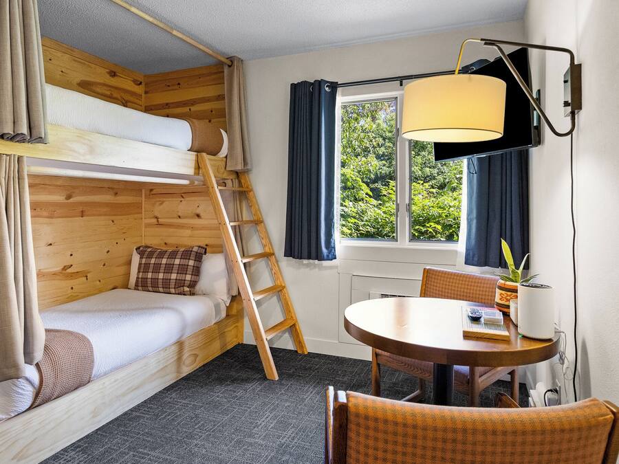 Quarto com beliche, mesa e cadeiras, luminária e janela com cortinas.