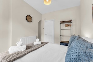 Quarto com cama, radiador, porta branca, prateleira de madeira com roupas e relógio de parede.