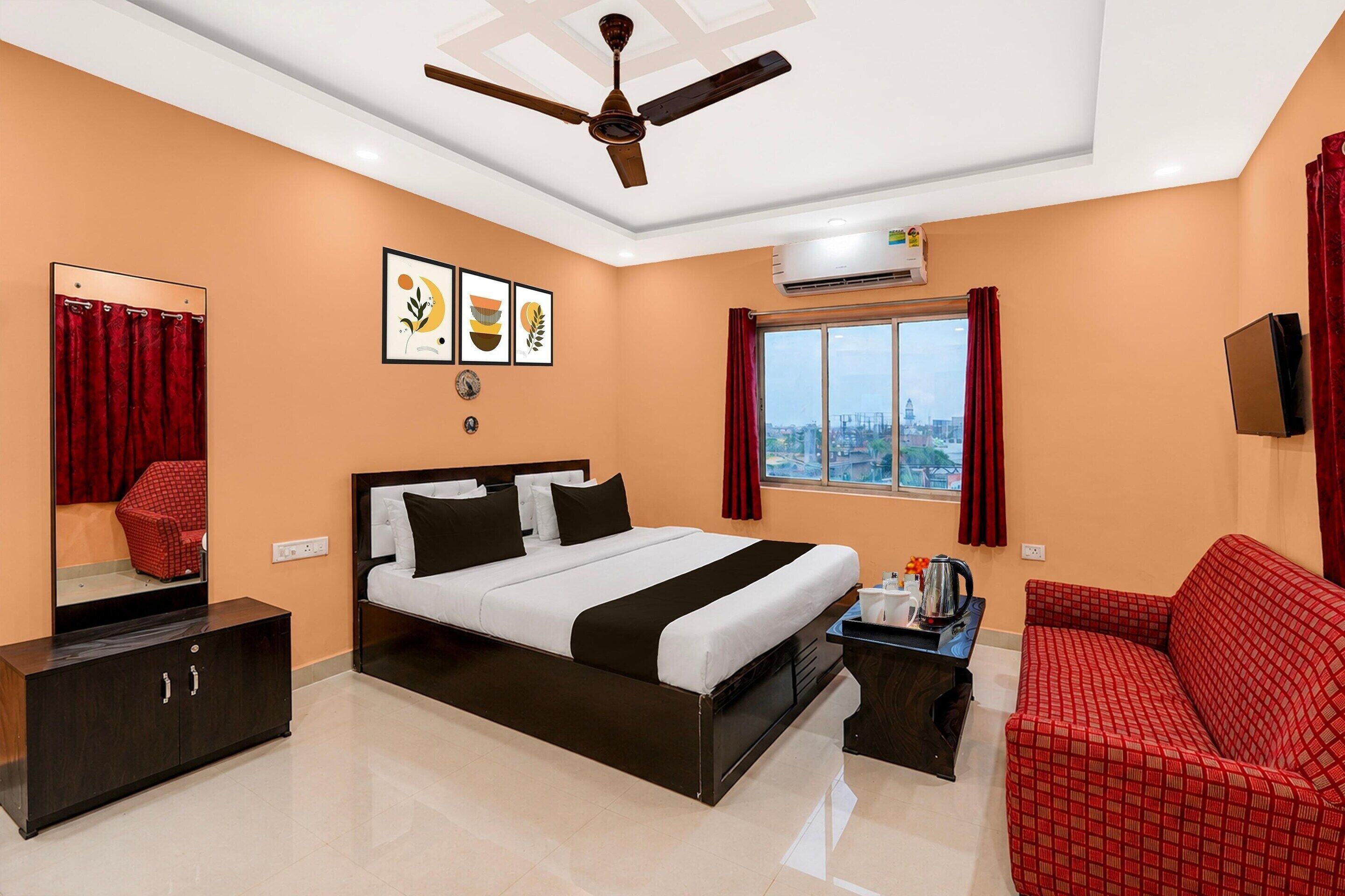 Quarto de hotel com cama, sofá, televisão, ventoinha no teto e janela com vista para a cidade.