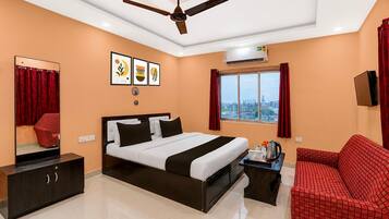 Quarto de hotel com cama, sofá, televisão, ventoinha no teto e janela com vista para a cidade.