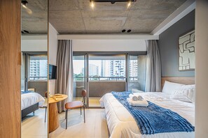 Quarto de hotel moderno com uma cama grande, uma escrivaninha, uma cadeira e vista para a cidade através de grandes janelas.
