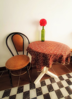 Uma área de refeições com uma mesa coberta por uma toalha estampada, uma cadeira com assento acolchoado e um vaso com flores vermelhas.