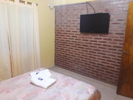 Quarto com parede de tijolos, televisão de tela plana fixada nela, cama com colcha estampada e uma porta à direita.