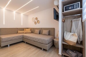 Um quarto moderno com um sofá-cama modular, uma mesa de cabeceira com abajur, um guarda-roupa com roupas penduradas e uma TV de tela plana na parede.