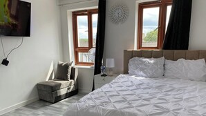 Um quarto com uma cama grande, uma cadeira, uma mesa de cabeceira com abajur e um relógio de parede.
