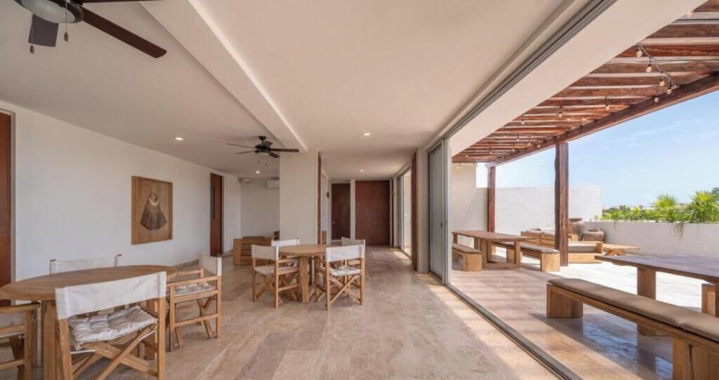Um espaço interior com uma longa mesa de jantar e bancos, um ventilador de teto e vista para um pátio exterior através de uma porta de vidro.