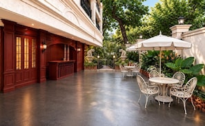 Corredor de hotel com área de bar, mesas externas com guarda-sóis e vista para o jardim.