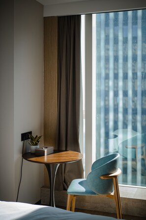 Quarto de hotel com uma cama, uma mesinha com uma planta, uma cadeira e vista para a cidade através de uma janela grande.