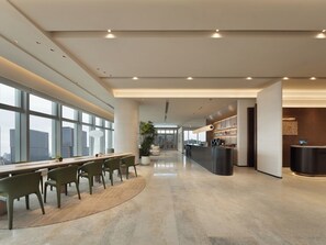 Um lobby de hotel moderno com um balcão de recepção extenso, área de estar com cadeiras, uma grande planta em vaso e vista para a paisagem urbana através de amplas janelas.