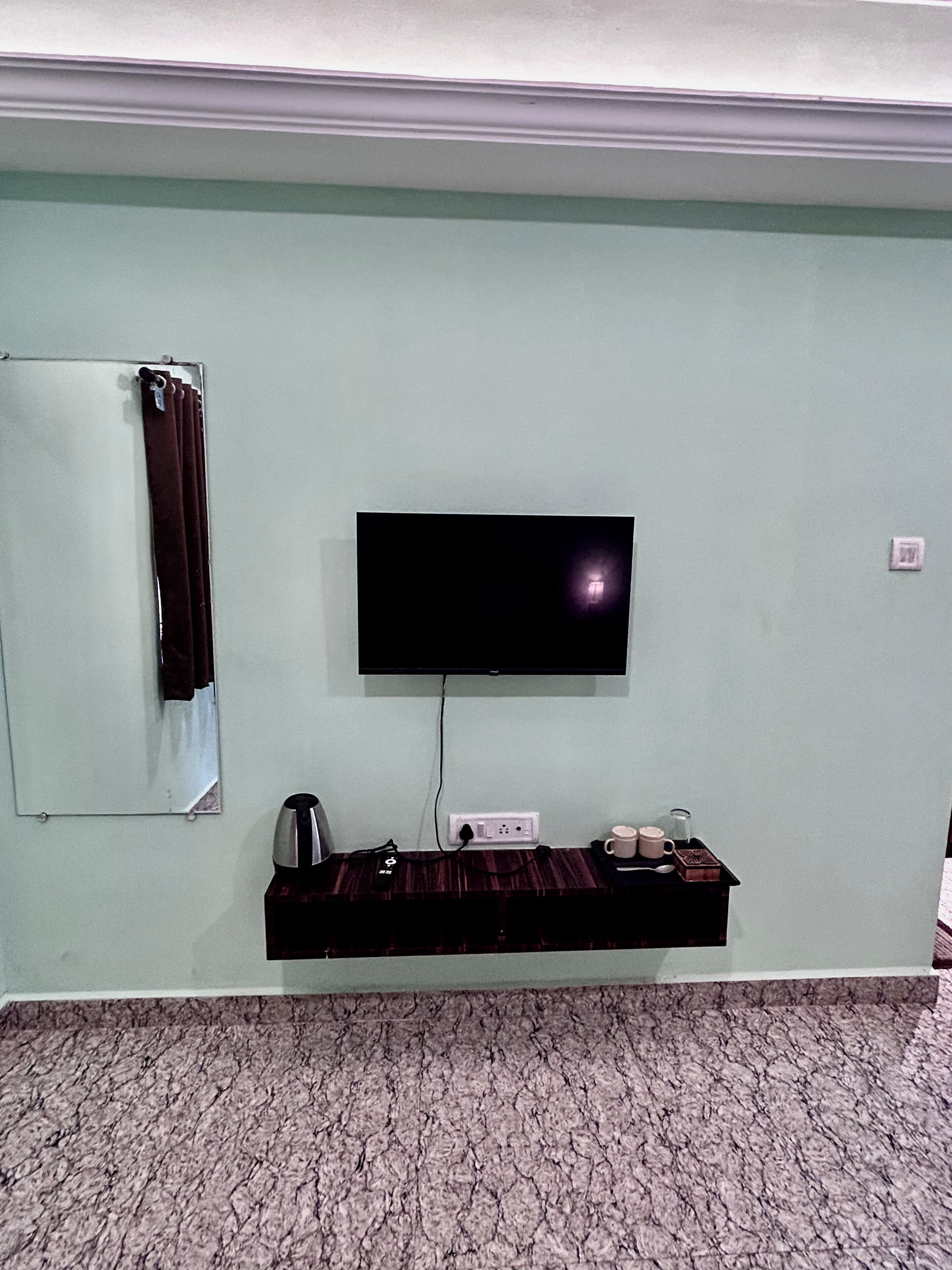 Quarto com televisão de tela plana na parede, uma pequena estante com chaleira e xícaras, e um tapete estampado.