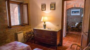 Quarto com cama, penteadeira, abajur e cadeira. Há uma janela e uma porta que dá para outro cômodo.