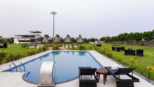 Piscina com espreguiçadeiras e uma mesa, cercada por vegetação e um edifício com várias janelas.