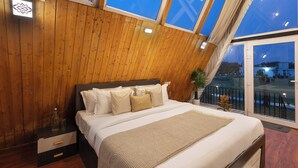 Quarto com teto inclinado de madeira, uma cama grande com várias almofadas, uma mesa de cabeceira e um vaso com planta.