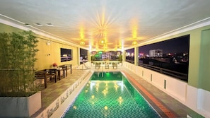Piscina no terraço com piso quadriculado, rodeada por assentos e mesas, com vista para o horizonte urbano à noite.