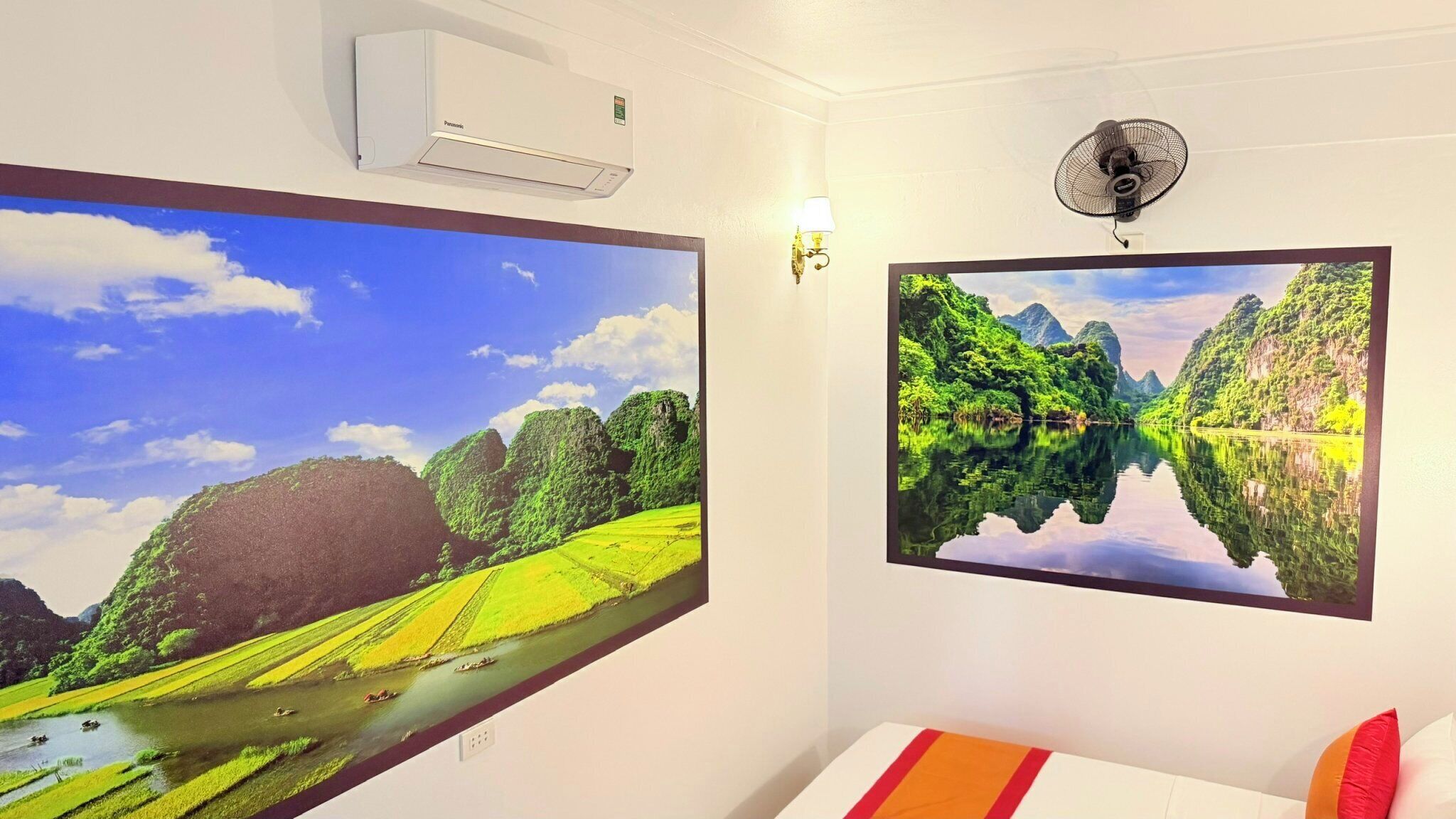 Quarto com duas grandes televisões fixadas na parede exibindo paisagens panorâmicas, um ventilador de teto e um ar-condicionado também fixado na parede.