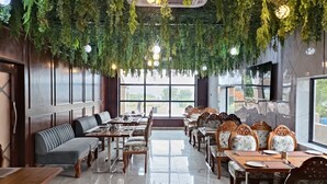 Restaurante com mesas e bancos grandes e compartilhados, teto alto com folhagem pendente e amplas janelas.