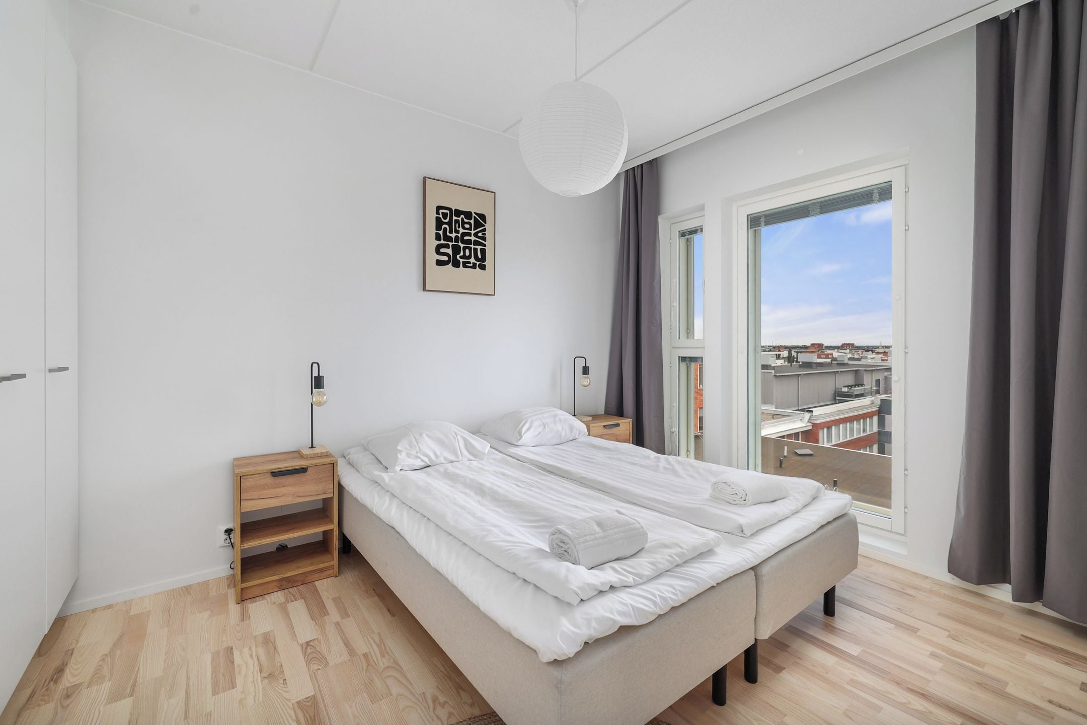 Quarto moderno com uma cama grande, mesas de cabeceira, luminária pendente e vista da cidade através de uma janela ampla.