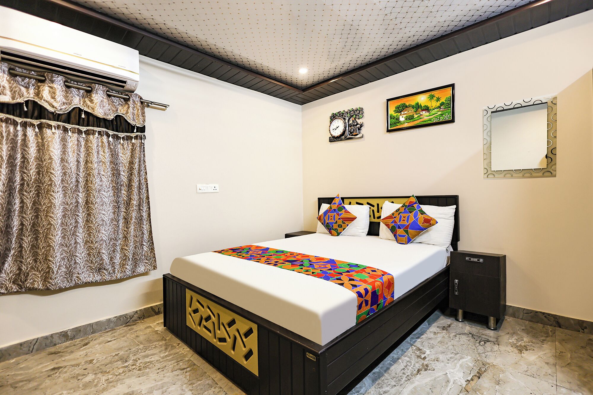 Quarto de hotel com cama, mesa de cabeceira, relógio de parede, quadro e espelho.