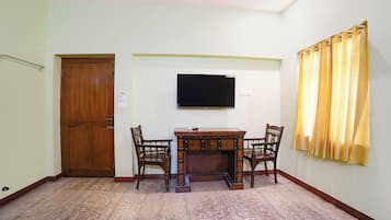Quarto com escrivaninha de madeira, duas cadeiras, televisão fixada na parede, porta e janela com cortinas.