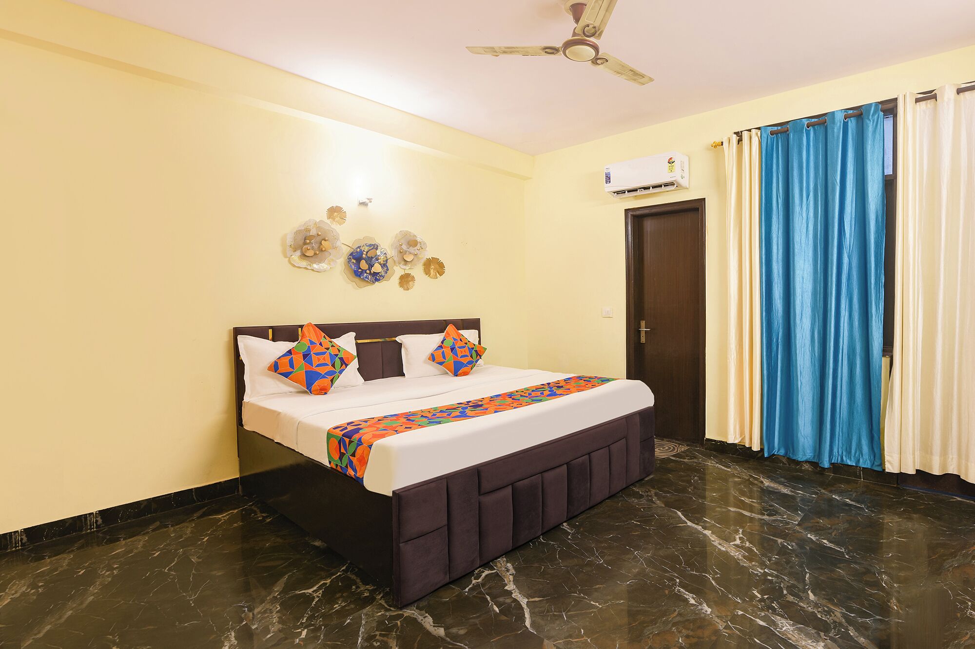 Quarto de hotel com cama, ventoinha no teto, ar condicionado, porta e janela com cortinas.