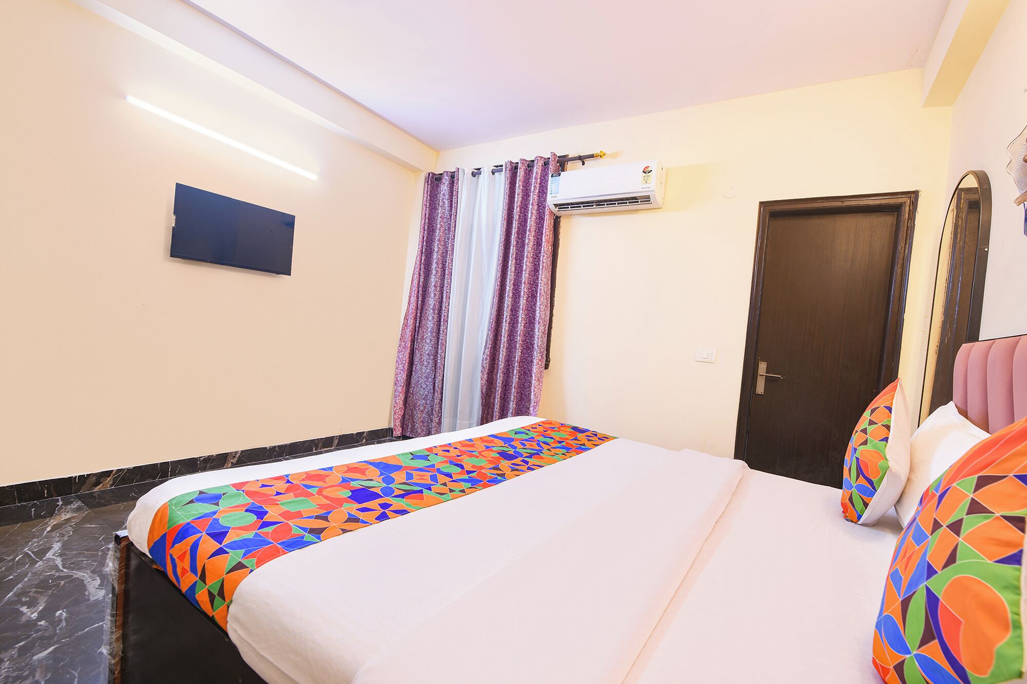 Quarto de hotel com cama, porta, ar-condicionado e televisão fixada na parede.