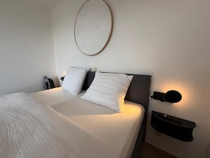Uma cama bem arrumada com lençóis brancos, um espelho redondo na parede e uma mesa de cabeceira com abajur.