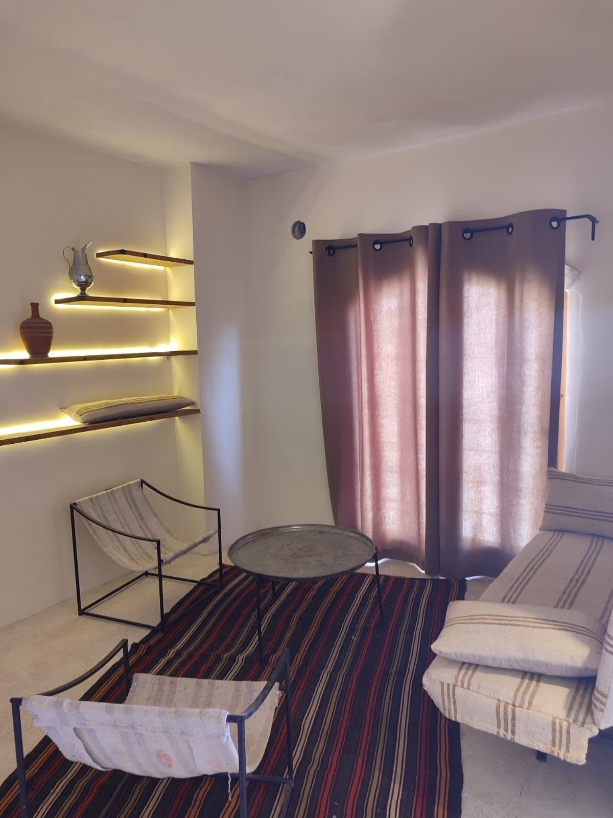 Um quarto com cama, uma cadeira, uma mesinha, uma estante com objetos decorativos e cortina.