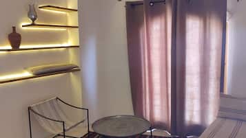 Um quarto com cama, uma cadeira, uma mesinha, uma estante com objetos decorativos e cortina.